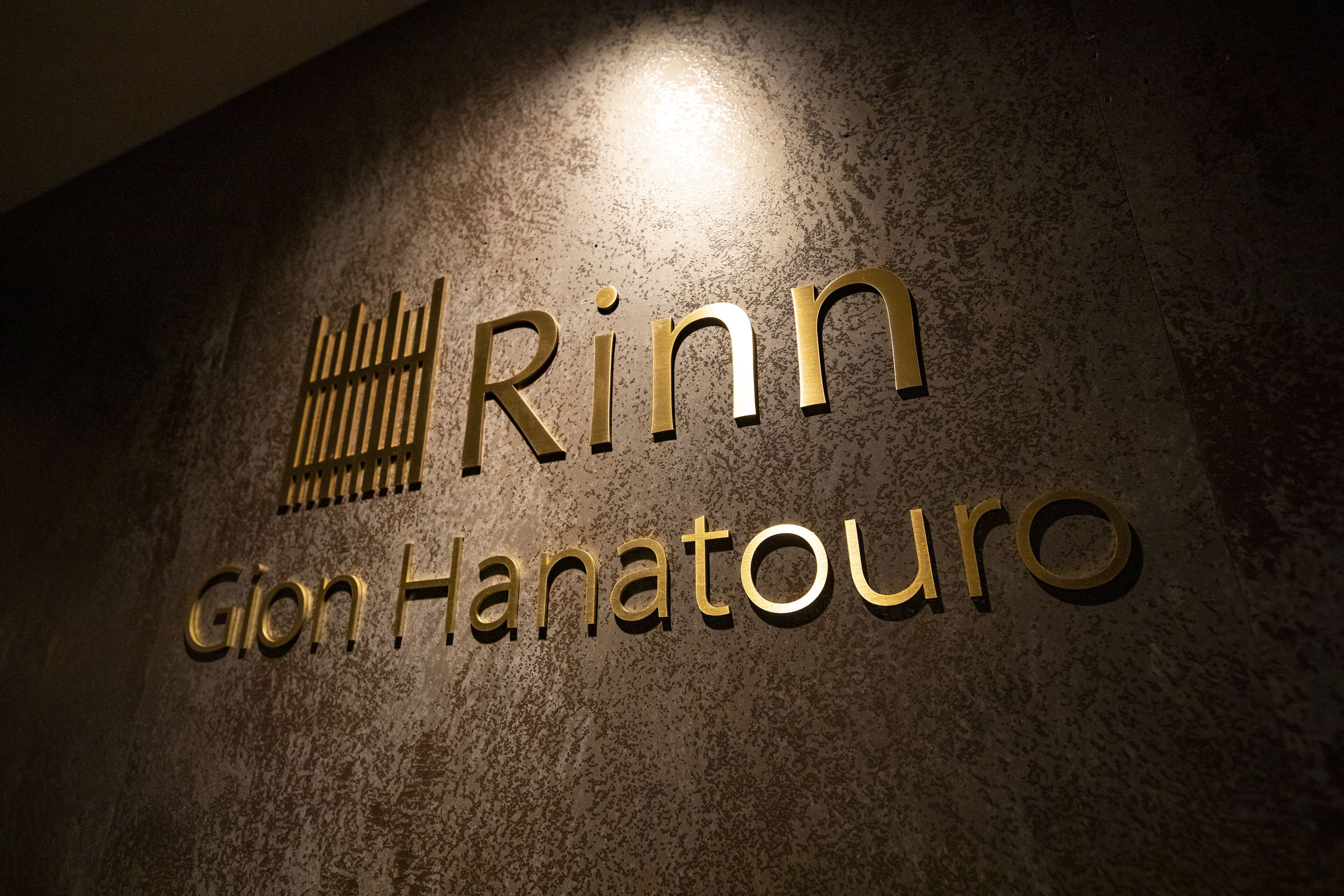 Um letreiro com os nomes 'Rinn' e 'Gion Hanatouro' em letras douradas sobre uma parede texturizada.