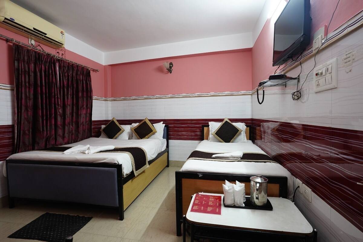 Quarto de hotel com duas camas, uma televisão, uma mesinha com um jarro e guardanapos, e uma cortina vermelha.