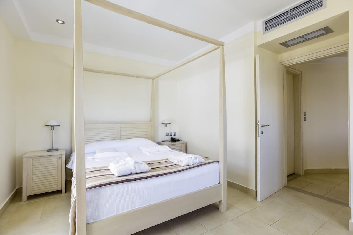 Uma cama de dossel com lençóis brancos, uma mesa de cabeceira com abajur e uma porta que dá para outro quarto.