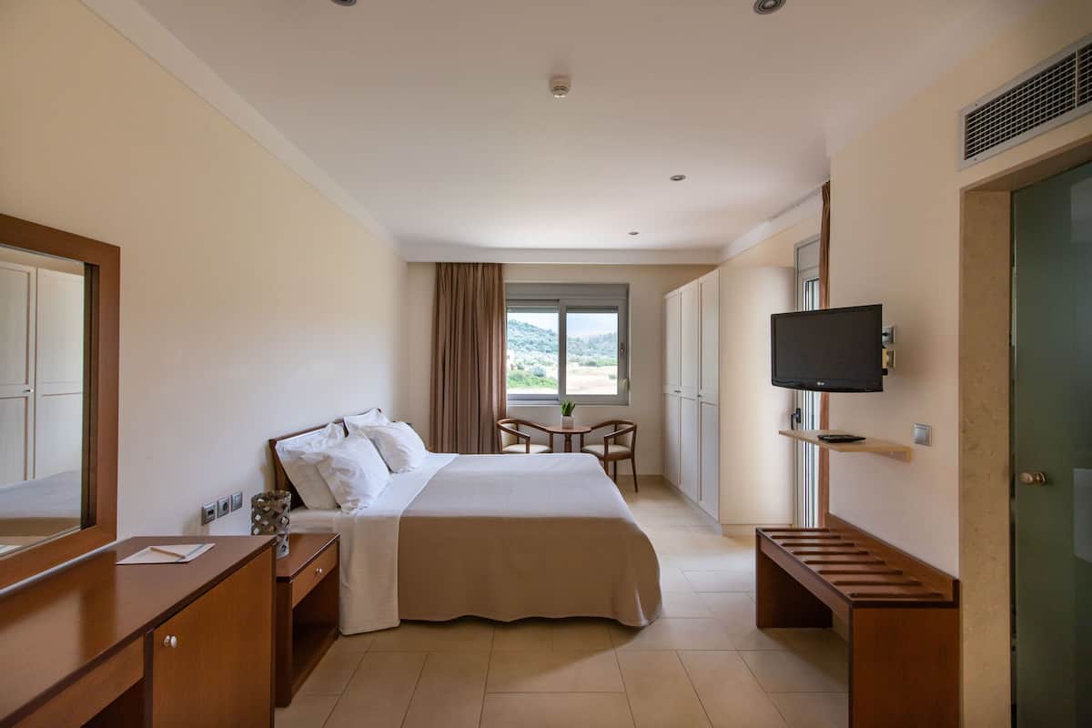 Quarto de hotel com cama, escrivaninha, cadeira, televisão e janela com cortinas.