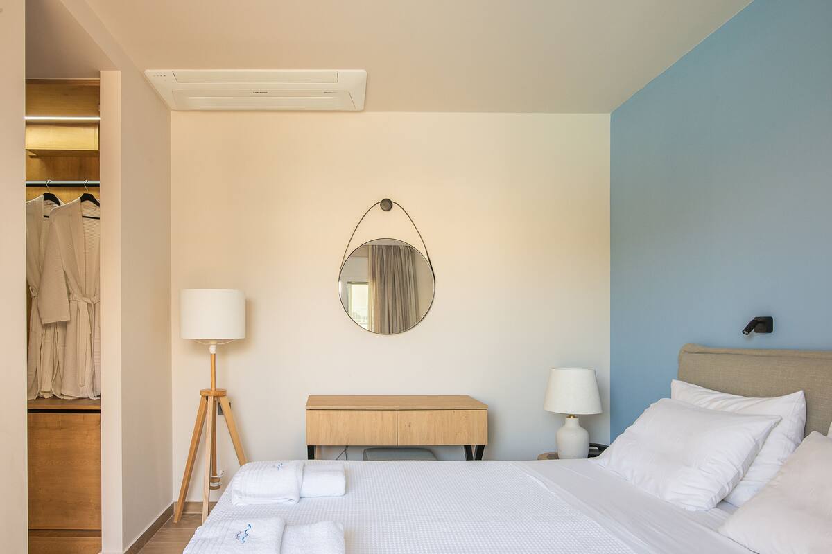 Quarto de hotel com cama, abajures, um espelho redondo, mesa de cabeceira de madeira e um guarda-roupa.
