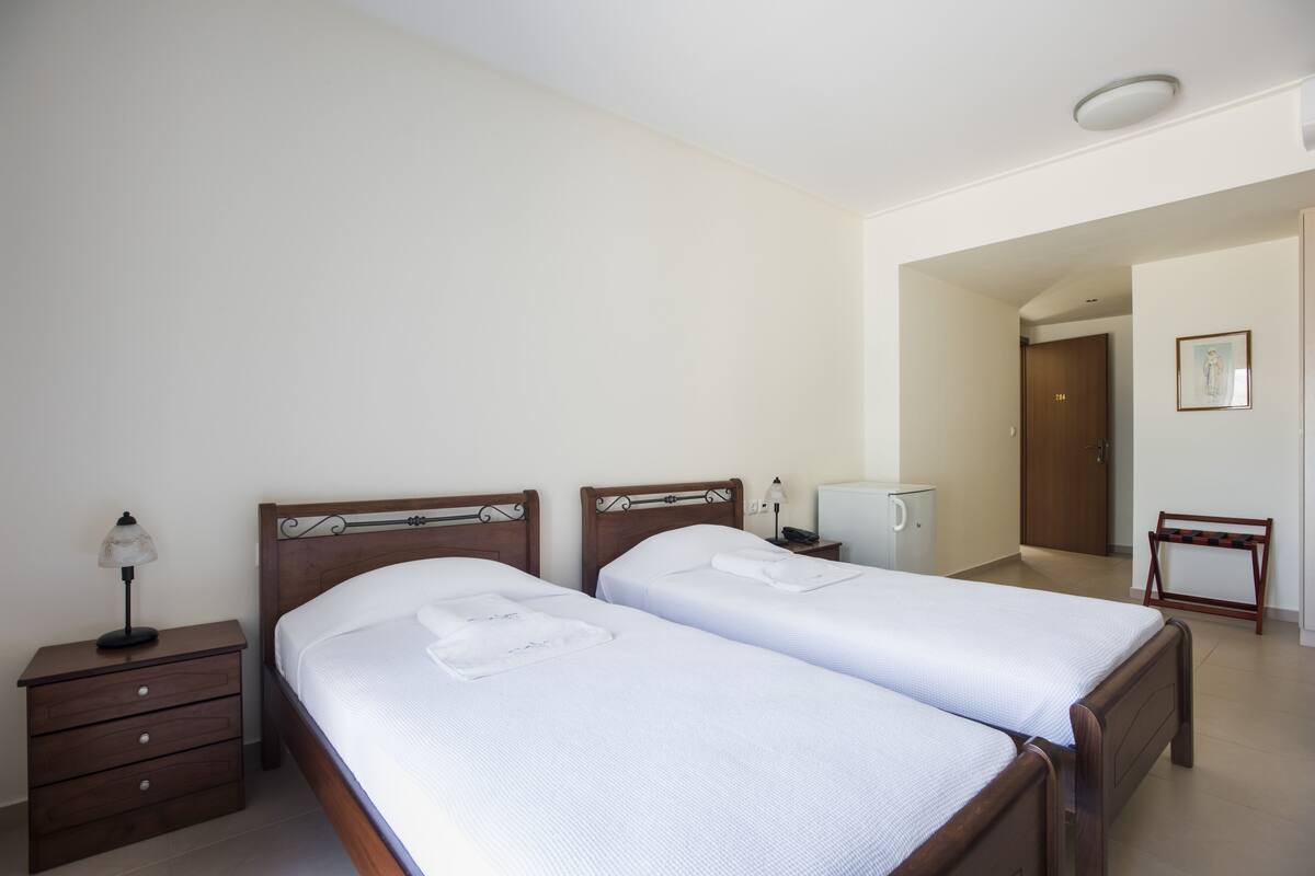 Quarto com duas camas de solteiro, mesa de cabeceira com abajur, pequeno armário e porta que dá para outro quarto.