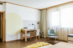 Quarto de hotel com escrivaninha de madeira, duas cadeiras, cama com cobertor amarelo, mesa pequena e janela com cortinas.