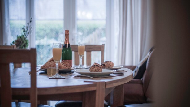 Uma mesa de jantar de madeira com croissants, uma garrafa de champanhe e copos de suco de laranja.