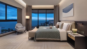 Quarto de hotel moderno com uma cama grande, uma cadeira, uma mesa de cabeceira e vista para o oceano.