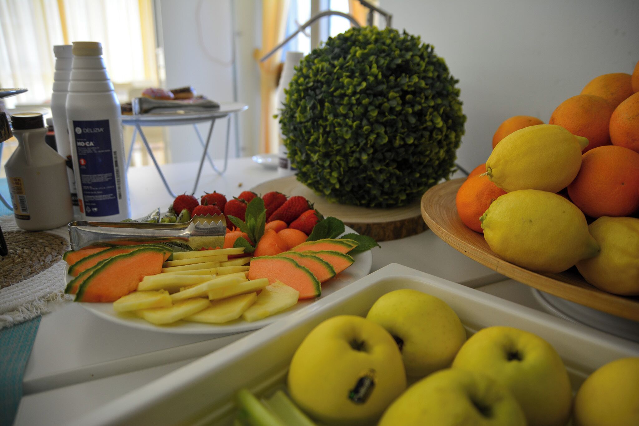 Uma mesa com diversas frutas e bebidas, incluindo morangos, maçãs, laranjas e uma planta decorativa.