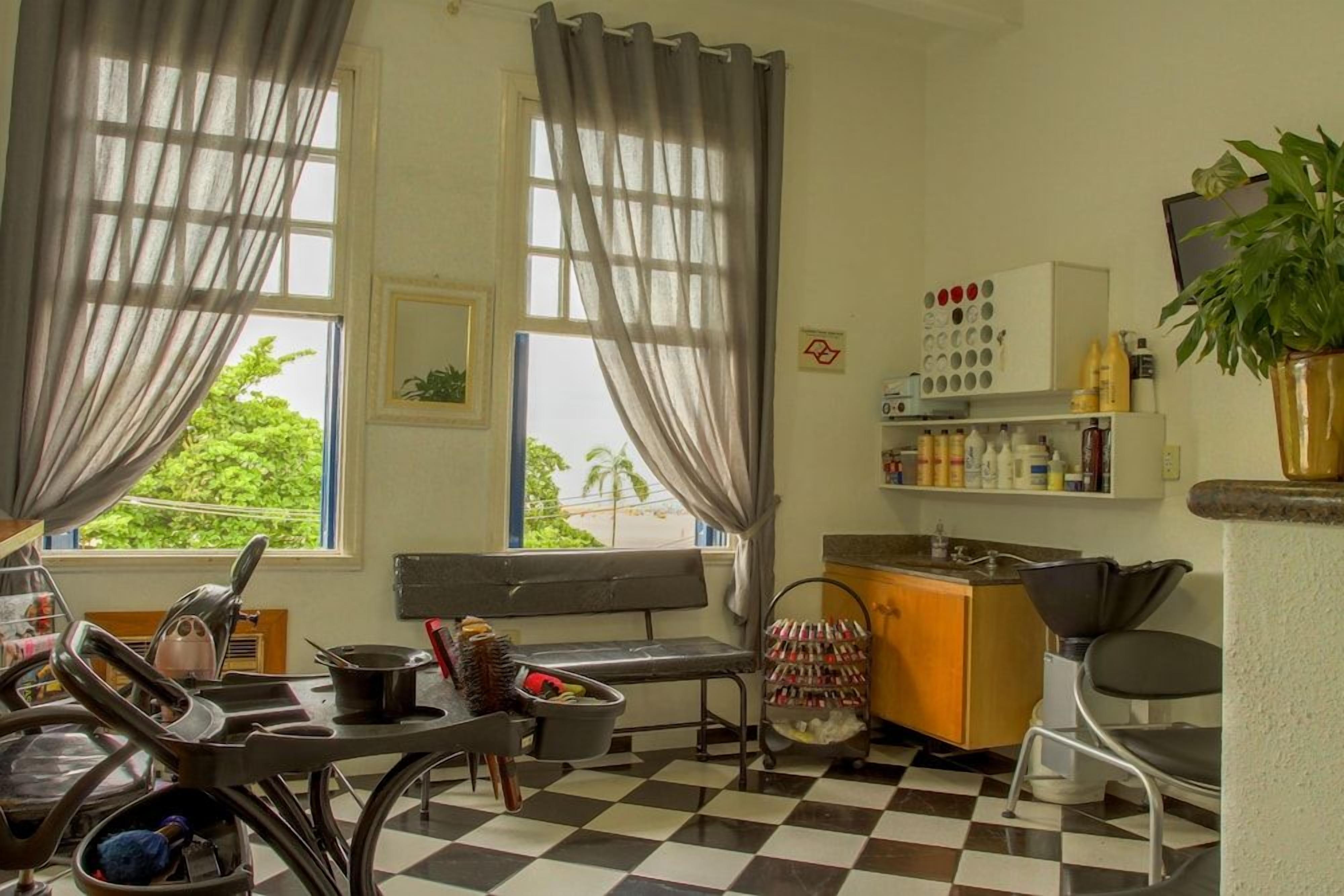 Um quarto com piso quadriculado, um banco, uma cadeira de cabeleireiro, uma pia e uma janela com cortinas.