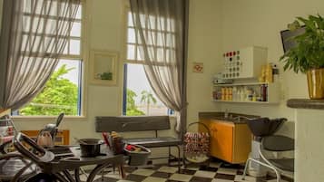Um quarto com piso quadriculado, um banco, uma cadeira de cabeleireiro, uma pia e uma janela com cortinas.