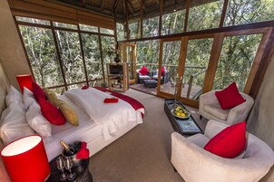 Quarto espaçoso com uma cama grande, área de estar com almofadas vermelhas e vista para a floresta através de janelas amplas.