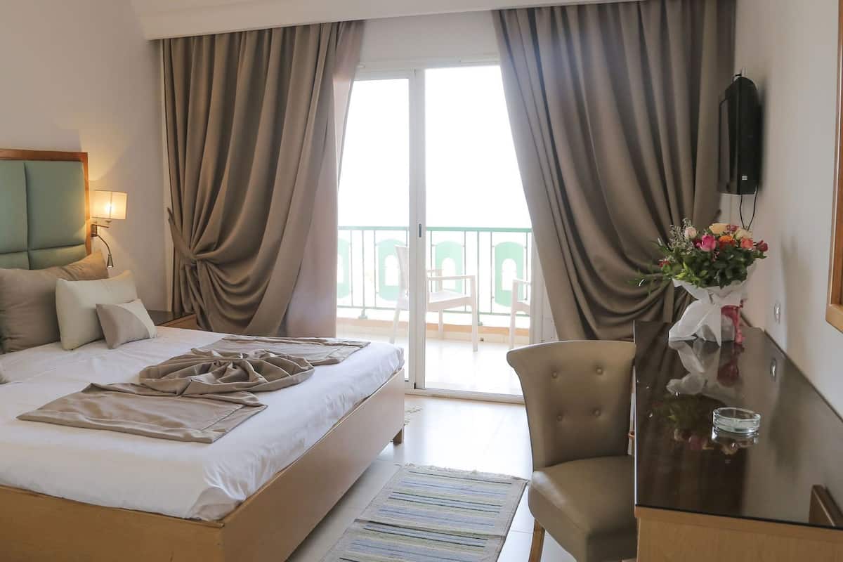 Quarto de hotel com cama, escrivaninha, cadeira, varanda com cortinas e televisão presa à parede.