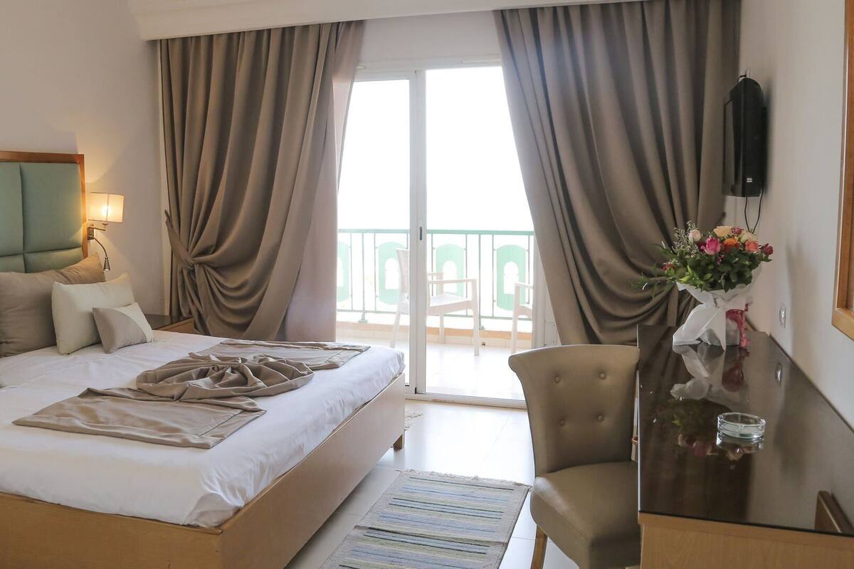 Quarto de hotel com cama, escrivaninha, cadeira, varanda com cortinas e televisão fixada na parede.