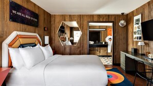 Quarto de hotel com uma cama grande, um espelho, uma mesa de trabalho, televisão e banheiro visível por uma porta aberta.