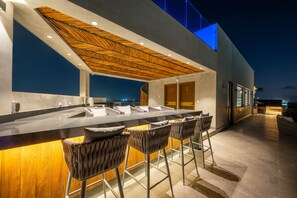 Área de bar ao ar livre com design moderno, apresentando teto de madeira, banquetas e um balcão comprido.