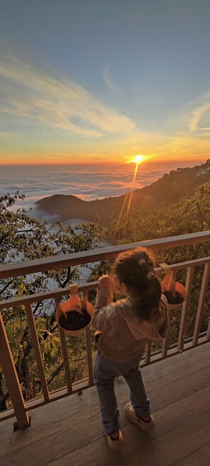 Criança em deck de madeira com vista para uma paisagem montanhosa ao pôr do sol.