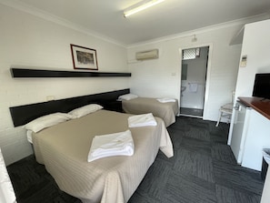 Quarto de hotel com duas camas, uma escrivaninha, uma cadeira, uma televisão e um banheiro visível através de uma porta aberta.