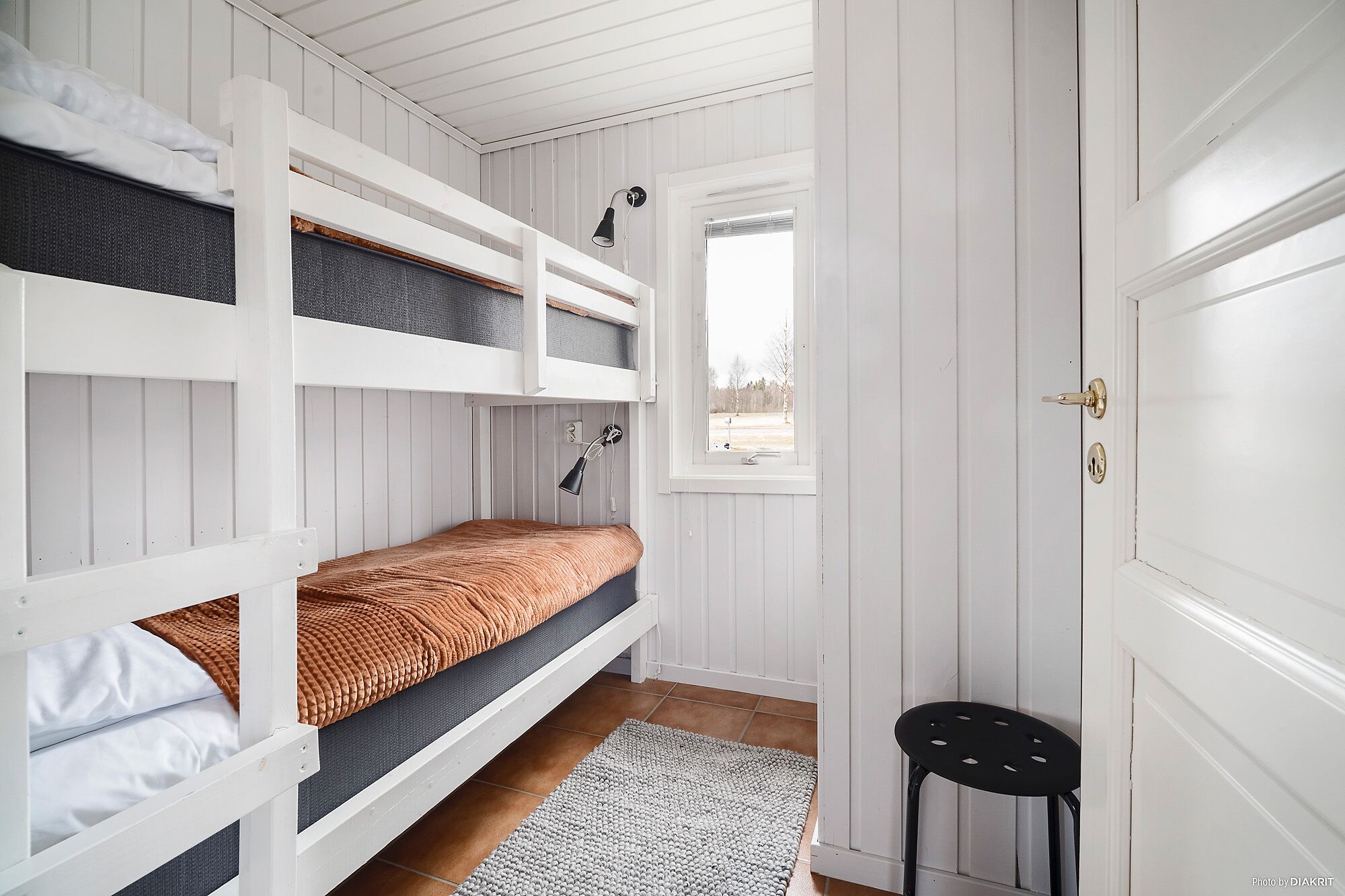 Quarto com beliche, janela, porta e um banquinho pequeno.