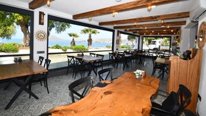 Restaurante com vigas de madeira, janelas amplas e vista para o mar. Mesas e cadeiras dispostas para refeições.