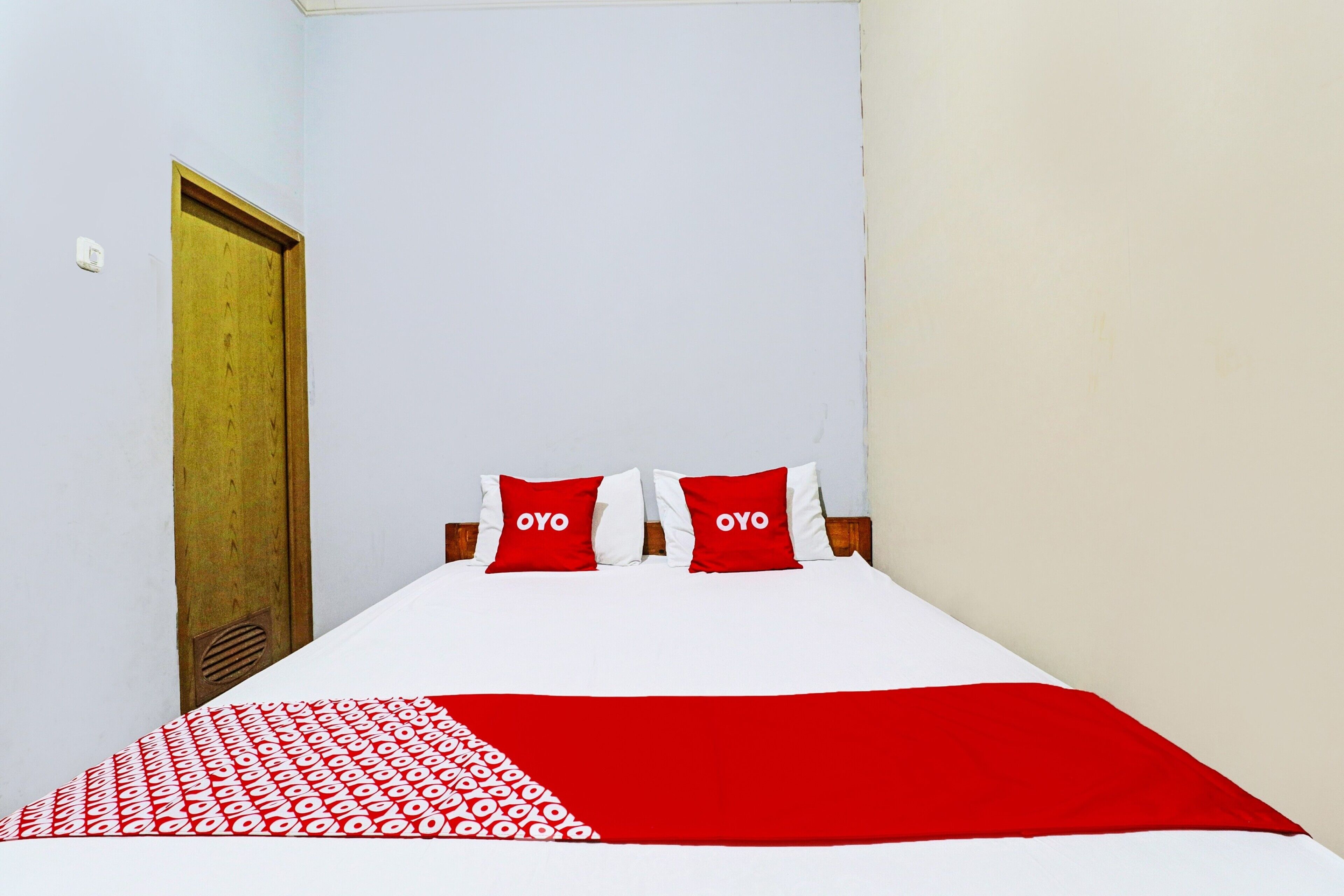 Quarto de hotel com uma cama, duas almofadas vermelhas com a palavra "OYO" estampada nelas, um cobreleito com padrão vermelho e branco e uma porta de madeira.