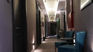 Corredor de hotel com piso em carpete, uma mesa com cadeiras e um extintor de incêndio na parede.