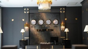 Uma área de recepção com uma parede escura, quatro relógios mostrando horários de Nova York, Londres, Istambul e Tóquio, e um candelabro acima.