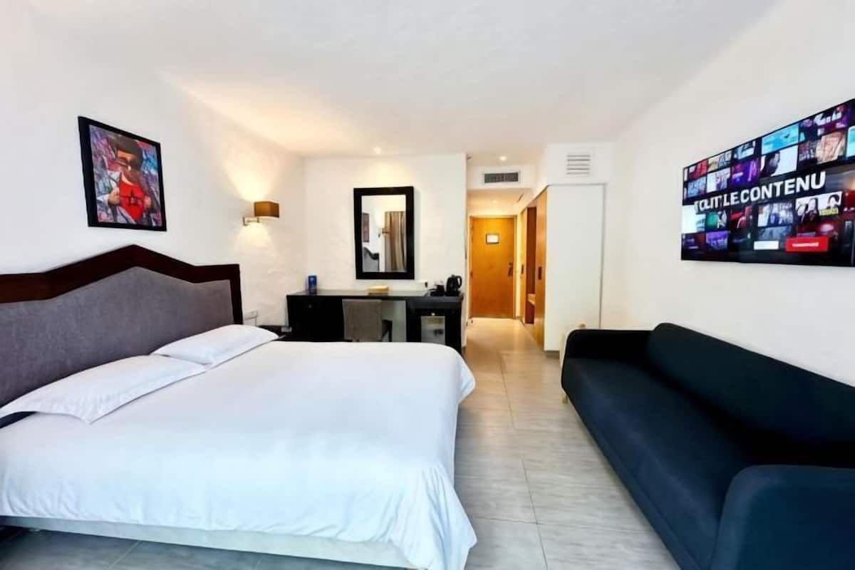 Quarto de hotel com cama, escrivaninha, cadeira, sofá, televisão e espelho.