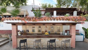 Uma área de bar ao ar livre com balcão de pedra, três banquetas e diversos frascos e copos.