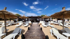 Uma área de lounge à beira-mar com cadeiras brancas, guarda-sóis de palha, um deck de madeira e um bar no centro.