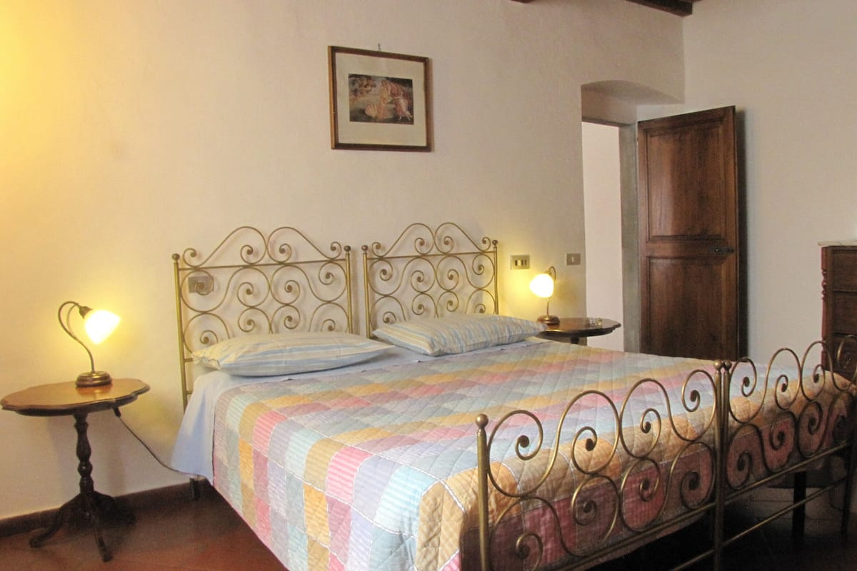 Quarto com estrutura de cama de metal, colcha acolchoada, duas mesas de cabeceira com abajures e porta de madeira.
