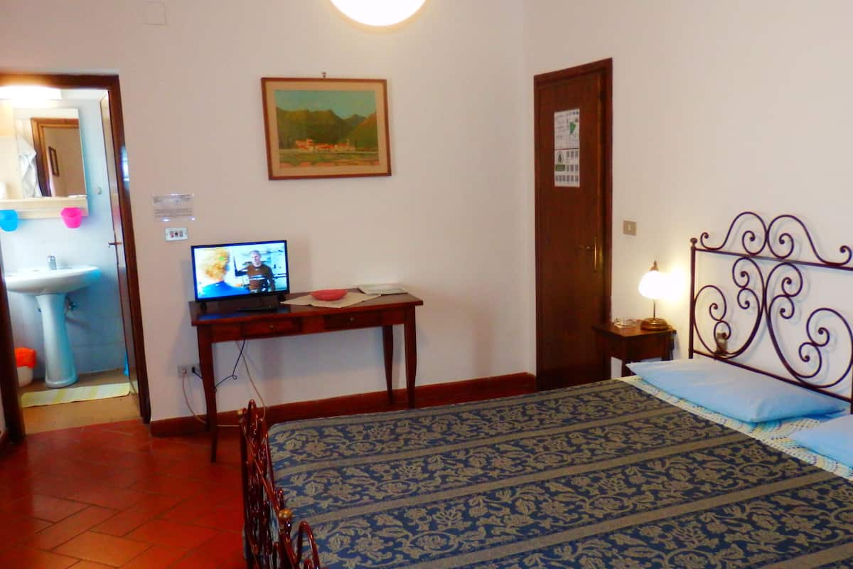 Quarto com cama, escrivaninha com computador, mesa de cabeceira com abajur, banheiro visível por uma porta aberta e um quadro na parede.