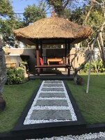 Um gazebo com telhado de palha, com um banco e uma mesa, cercado por um caminho de pedra e vegetação.