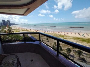 Varanda com vista para uma praia, palmeiras e um horizonte urbano ao longe.