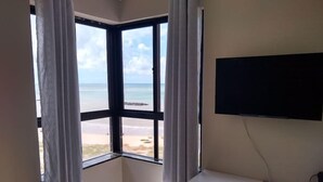 Quarto com uma grande janela com vista para a praia, uma televisão de tela plana na parede e uma pequena escrivaninha.