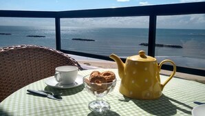 Uma mesa com toalha xadrez, um bule, uma xícara e um copo de sobremesa junto a uma janela com vista para o mar.