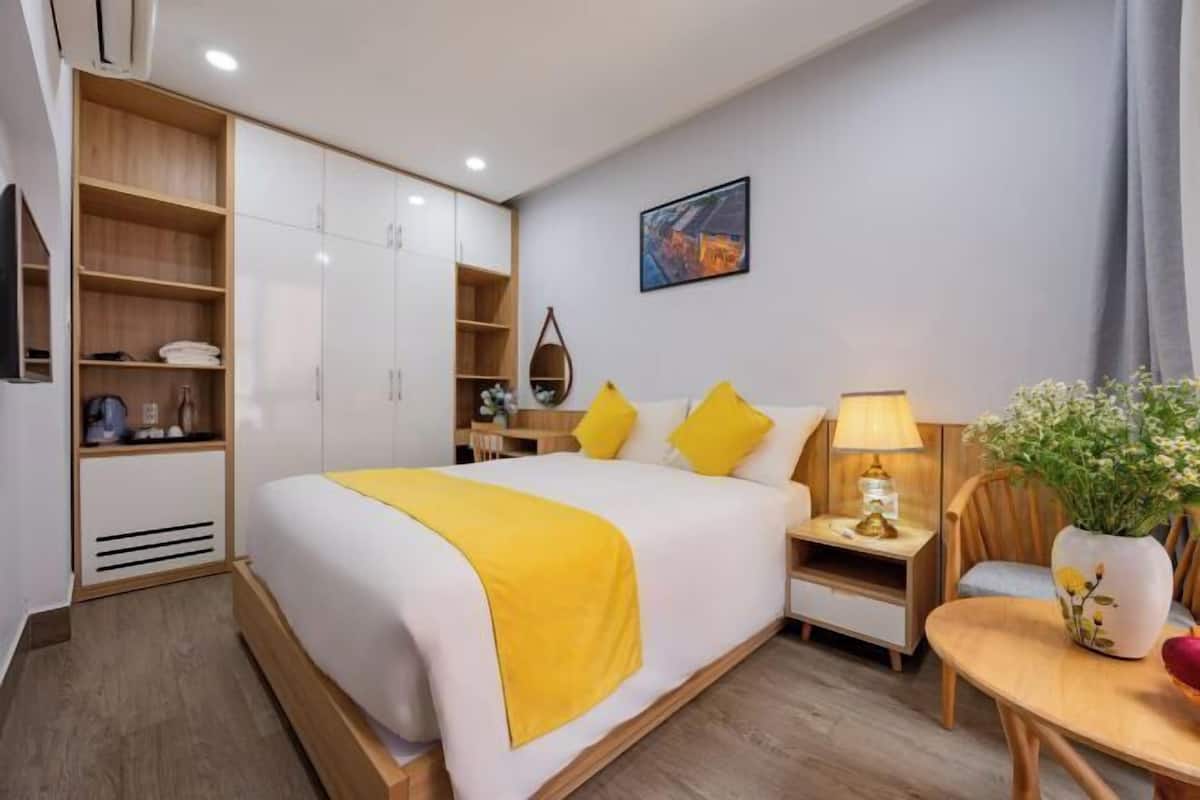Quarto de hotel com cama, mesa de cabeceira com abajur, poltrona, pequena mesa com um vaso e guarda-roupa.