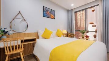 Quarto de hotel com cama, escrivaninha, cadeira, uma janela grande e luminária pendente.