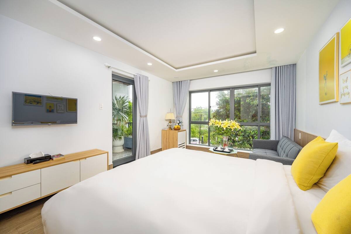 Quarto de hotel moderno com uma cama grande, televisão na parede, janela com cortinas e uma pequena mesa com abajur.