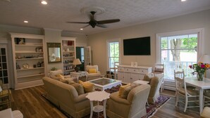 Uma sala de estar ampla com ventilador de teto, estantes embutidas, uma janela grande e um espaço de jantar com mesa e cadeiras.