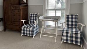 Um quarto com um armário de madeira, duas poltronas xadrez, uma mesa pequena e uma janela com persianas.