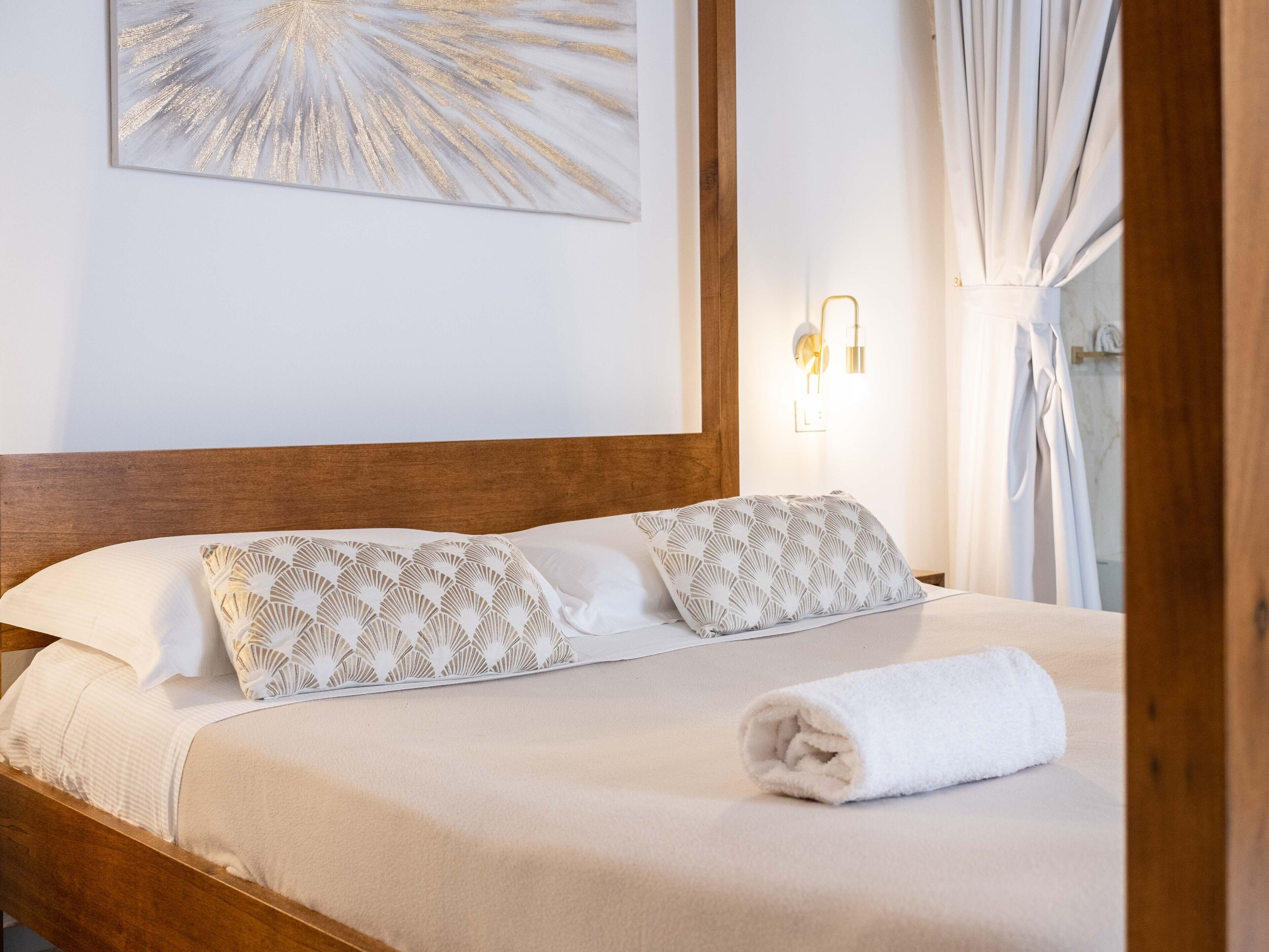 Cama bem arrumada com lençóis brancos e almofadas estampadas, cabeceira de madeira, luminária fixada na parede e uma obra de arte abstrata emoldurada acima da cama.