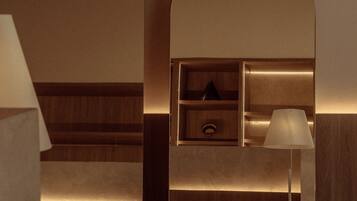 Quarto de hotel moderno com piso de madeira, sofá com almofadas listradas, mesa de cabeceira com abajur e prateleira embutida com luminária.