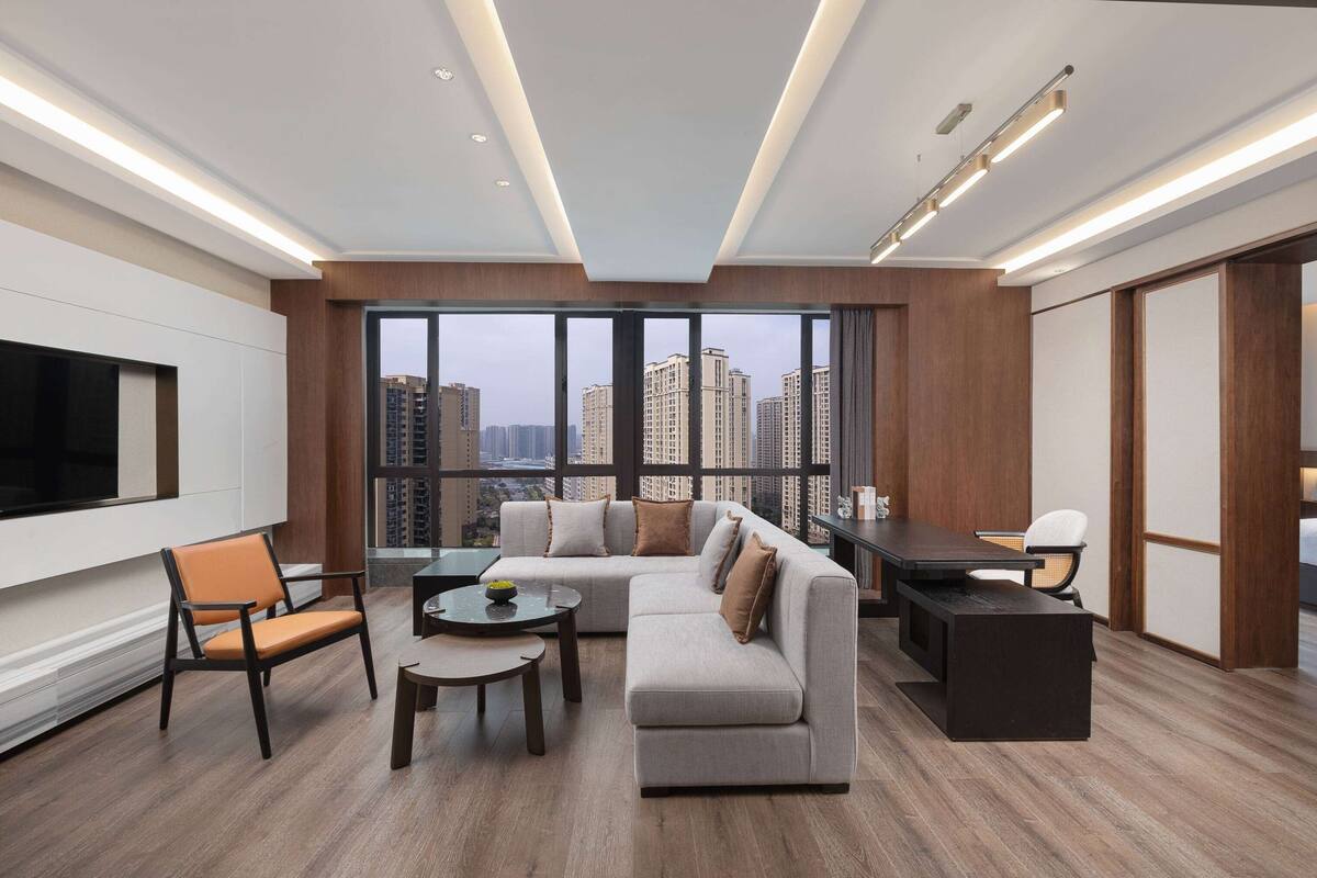 Uma sala de estar moderna com sofá seccional, uma mesa de centro, televisão fixada na parede e grandes janelas com vista para a cidade.