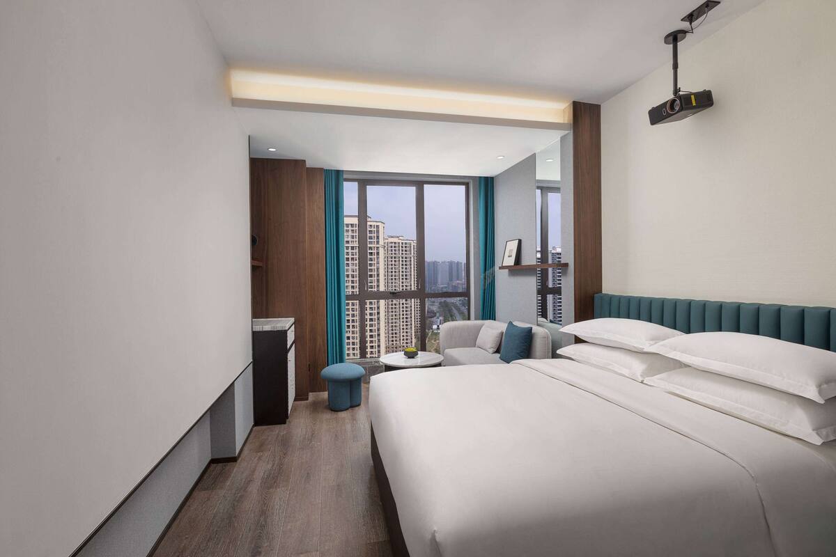 Quarto de hotel moderno com uma cama grande, uma mesinha, um sofá e vista para edifícios altos.