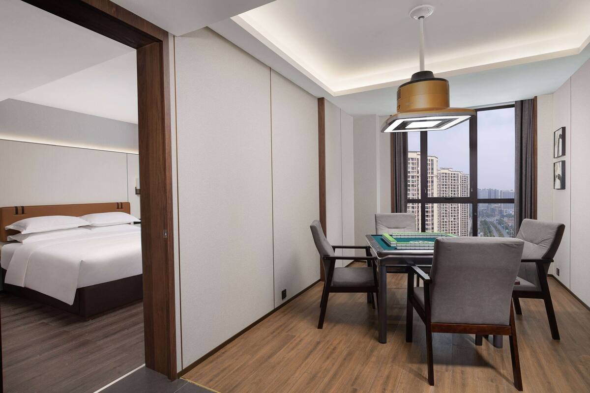 Quarto de hotel com cama, mesa de jantar com cadeiras, luminária pendente e uma grande janela com vista para a cidade.