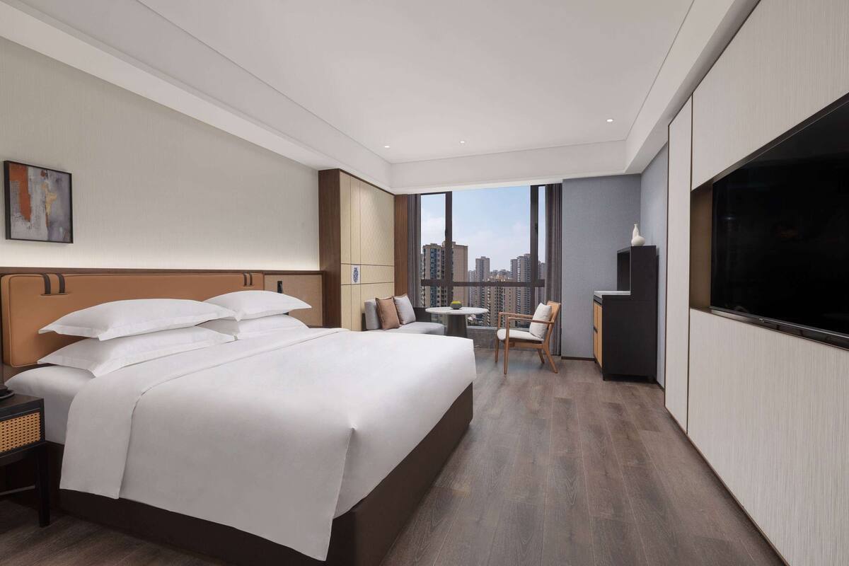 Quarto de hotel moderno com uma cama grande, uma escrivaninha, uma cadeira e vista para o horizonte da cidade.