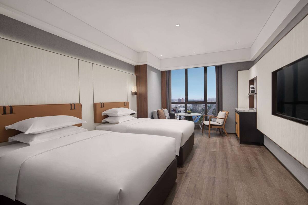 Quarto de hotel com duas camas, uma escrivaninha e uma grande janela com vista para a cidade.