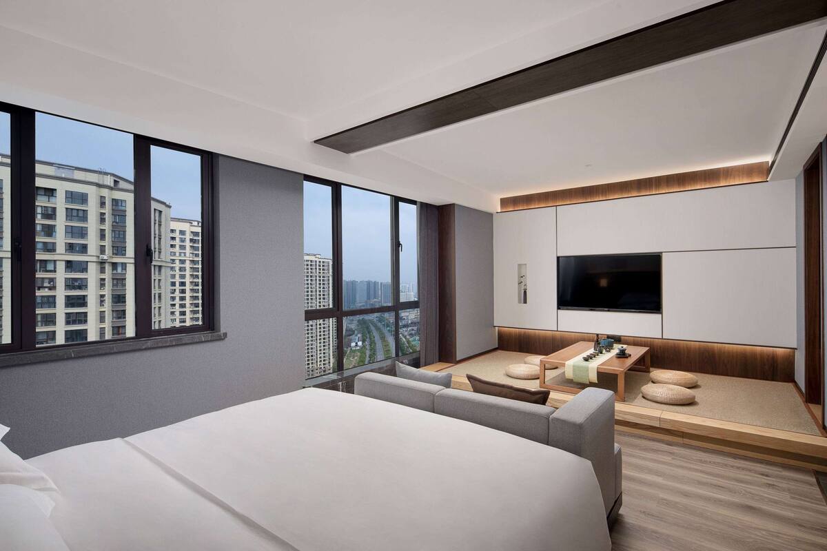 Quarto de hotel moderno com uma cama grande, área de estar com TV e vista para edifícios altos.