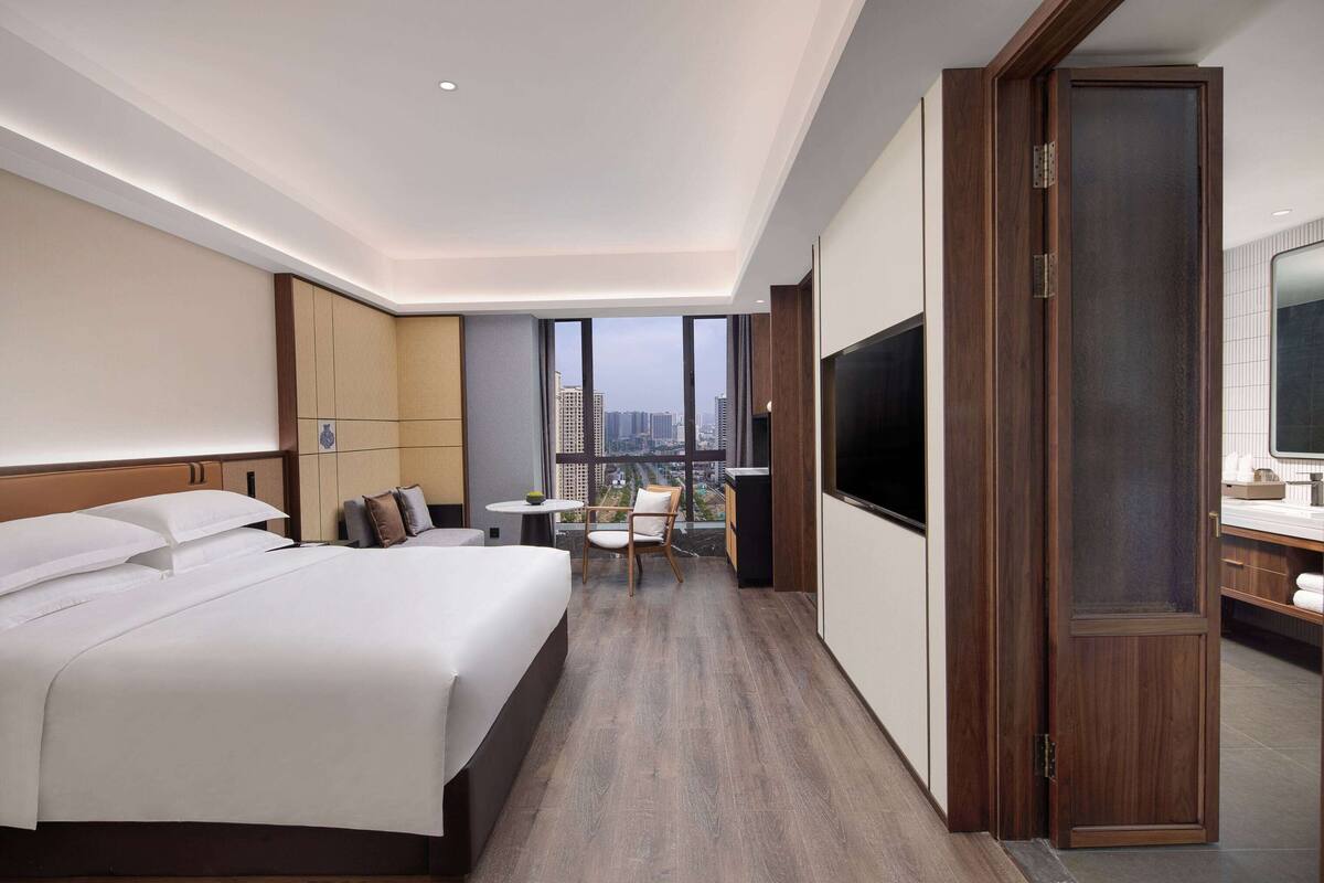 Quarto de hotel moderno com uma cama grande, televisão, varanda com vista para a cidade e uma pequena mesa com cadeiras.