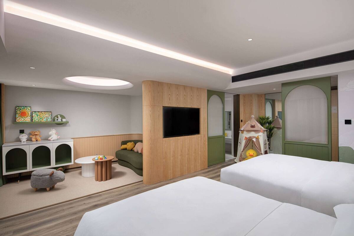 Quarto de hotel moderno com televisão de tela plana, sofá, mesa redonda pequena e duas camas.