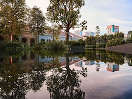 Um corpo d'água calma refletindo edifícios e árvores, rodeado por vegetação.