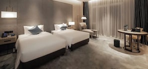 Quarto de hotel com duas camas, uma mesa pequena, uma cadeira, um abajur e uma janela grande com cortinas.